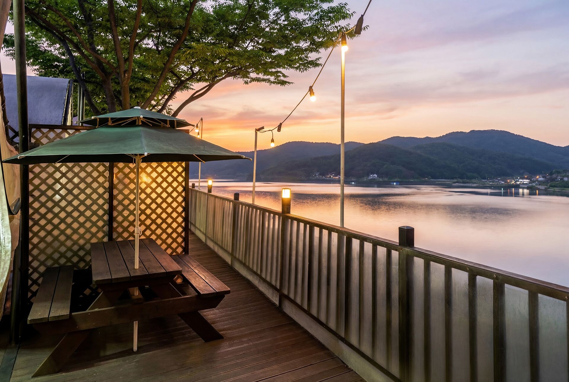 Um deck de madeira com mesa de piquenique e área de estar coberta, com vista para águas calmas e montanhas ao longe.