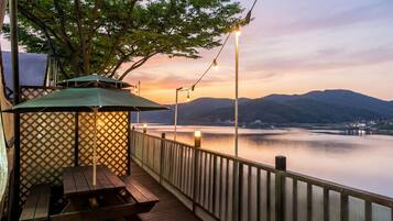 Um deck de madeira com mesa de piquenique e área de estar coberta, com vista para águas calmas e montanhas ao longe.