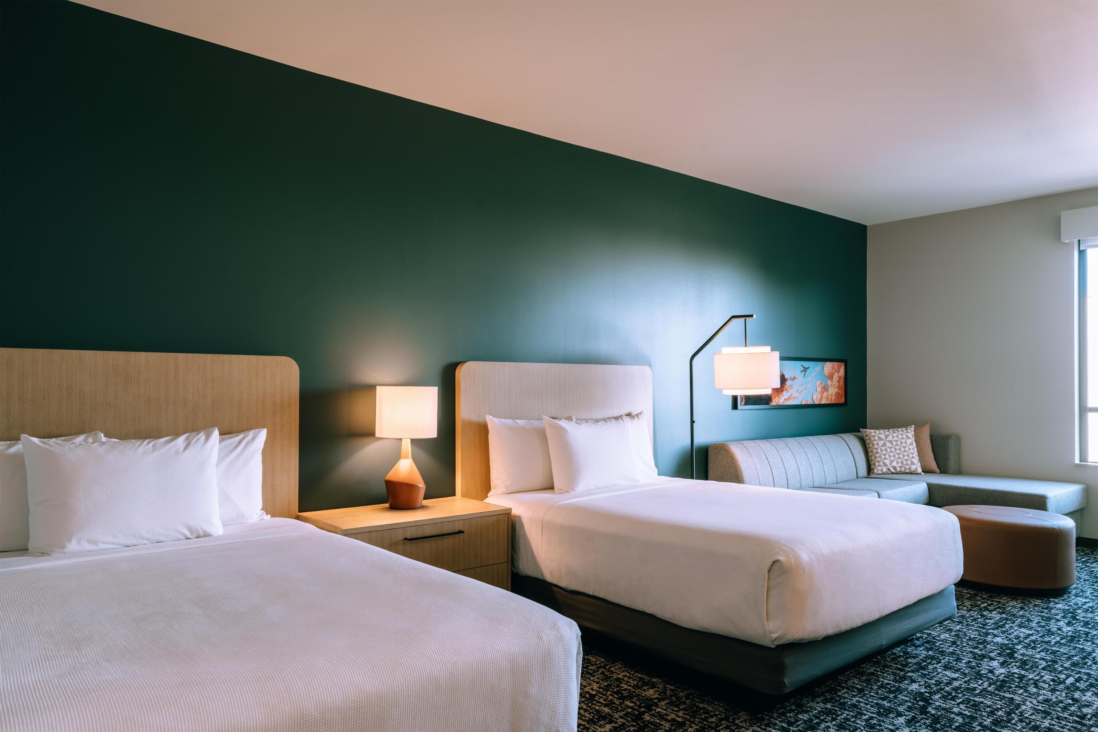 Quarto de hotel com duas camas, uma escrivaninha, luminária, sofá e um quadro na parede.