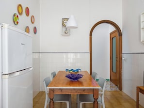 Uma cozinha com mesa de madeira, cadeiras brancas, frigorífico e pratos decorativos na parede.