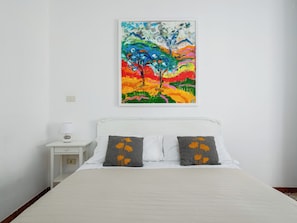 Uma cama com roupa de cama branca e duas almofadas, uma mesa de cabeceira com abajur e um quadro abstrato colorido na parede.