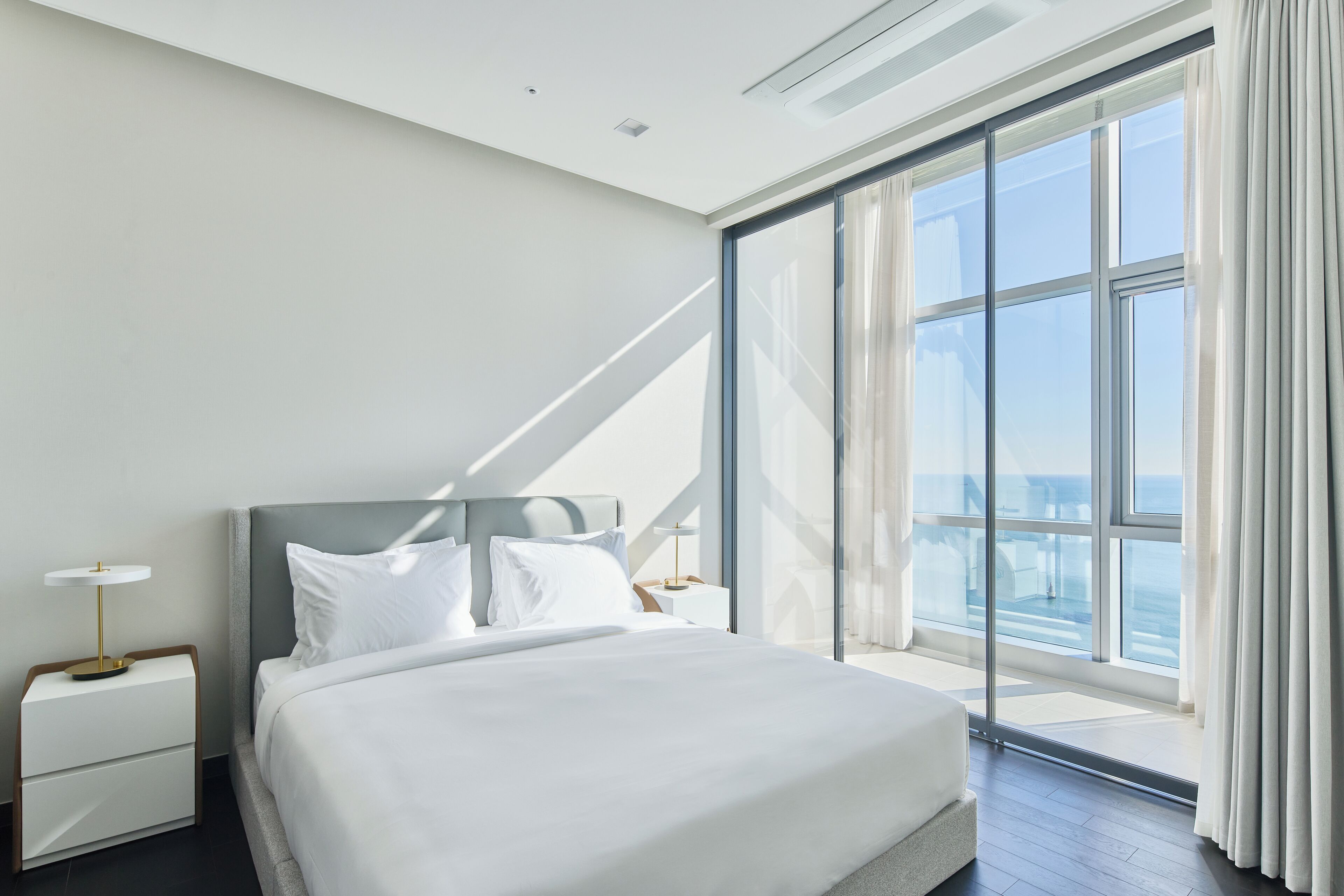 Quarto de hotel moderno com uma cama grande, mesa de cabeceira e vista do oceano através de janelas do chão ao teto.