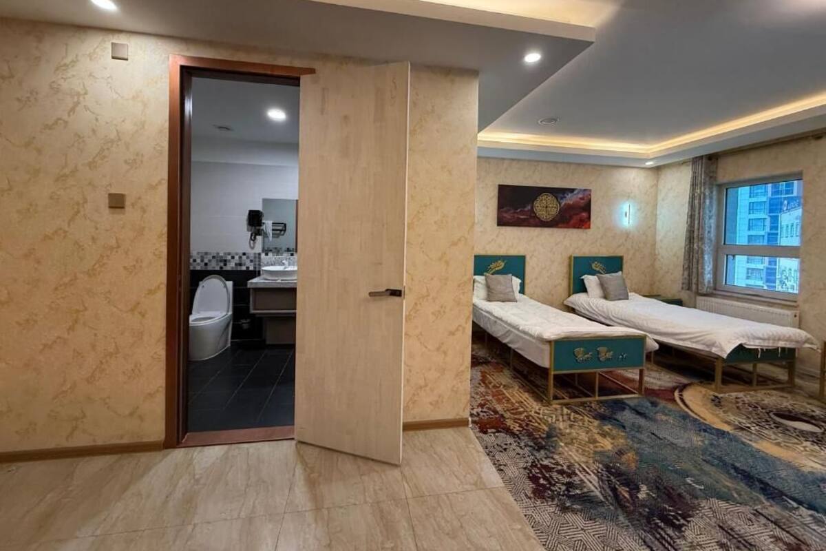Quarto de hotel com duas camas de solteiro, banheiro com vaso sanitário e pia, uma janela grande e um tapete estampado.