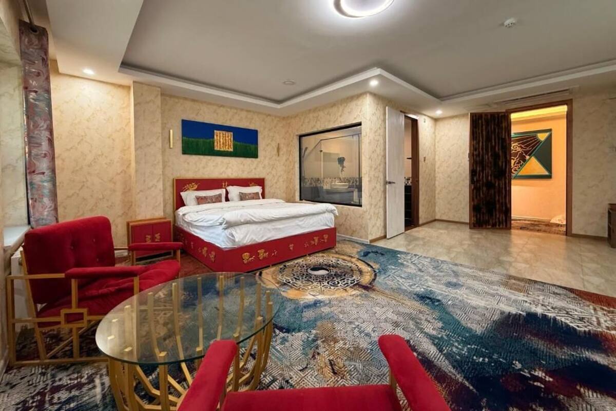 Quarto de hotel com uma cama grande, uma mesa de centro de vidro, cadeiras vermelhas e um tapete estampado.