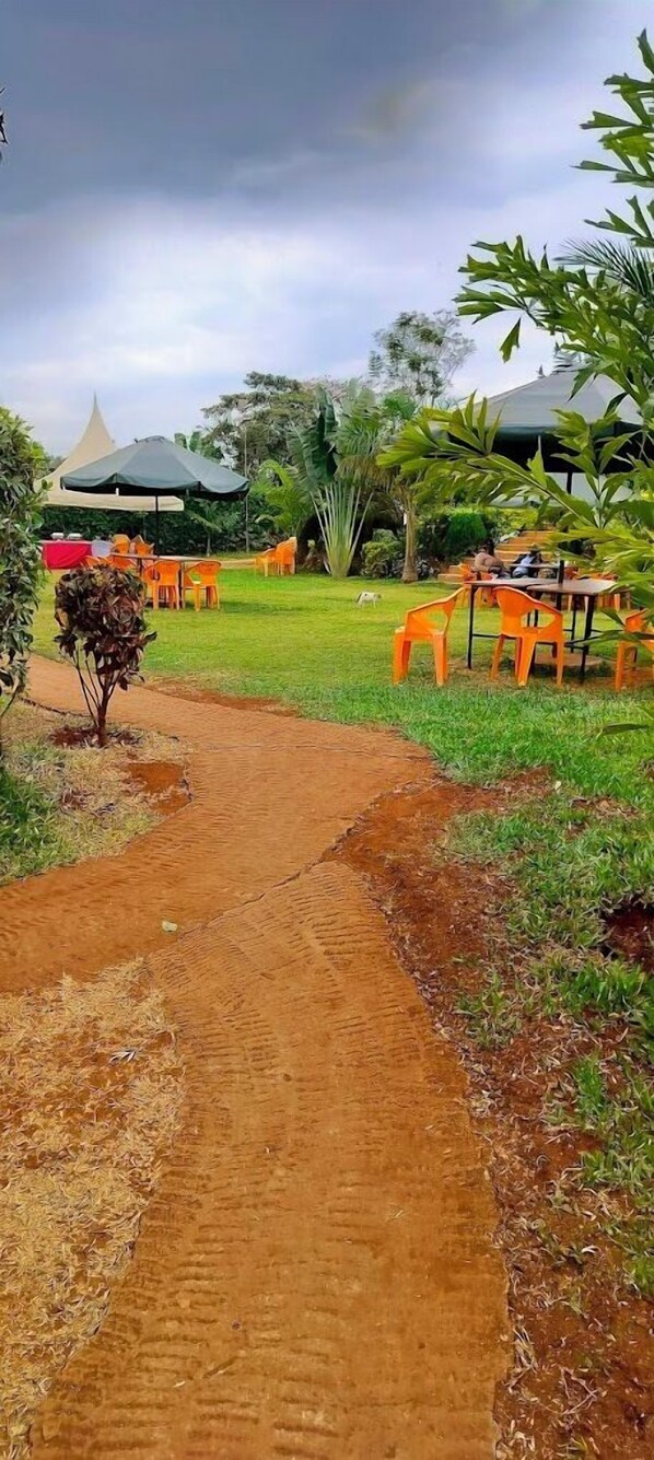 Uma trilha de terra leva a uma área de estar com cadeiras laranja e uma mesa, tudo cercado por vegetação e uma tenda.