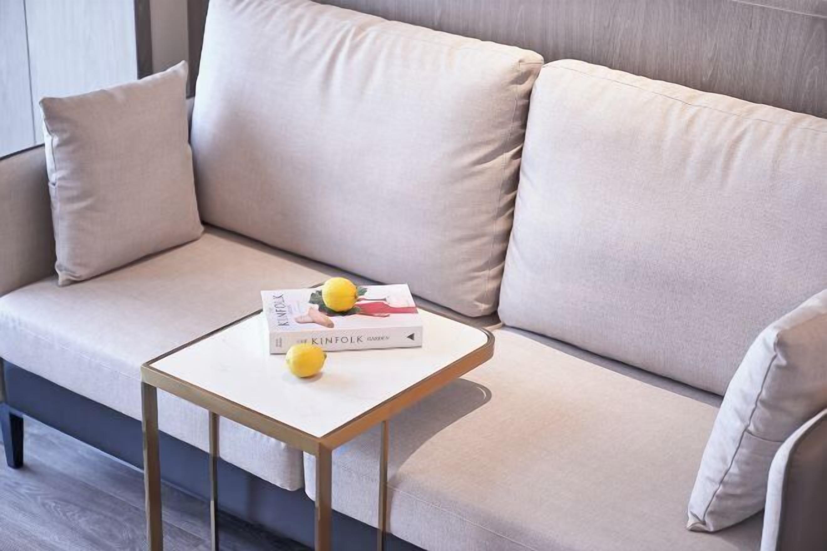 Um sofá com almofadas, uma pequena mesa com um livro e um objeto amarelo, e uma janela ao fundo.