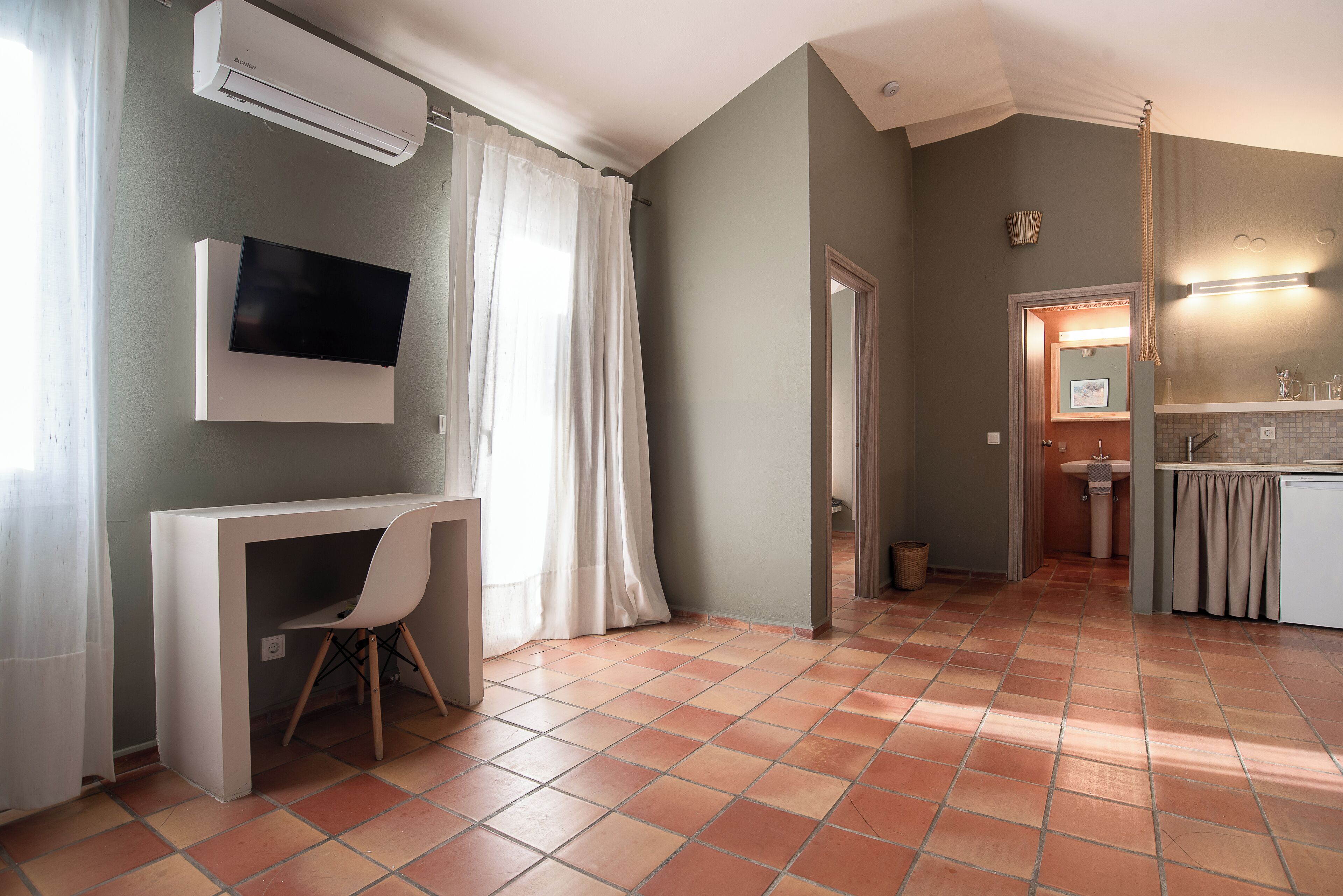 Quarto com escrivaninha, cadeira, ar condicionado e vista para um banheiro.
