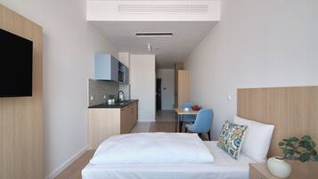 Quarto de hotel moderno com cama, mesa de cabeceira, pequena escrivaninha com cadeira, cozinha compacta e TV fixada na parede.