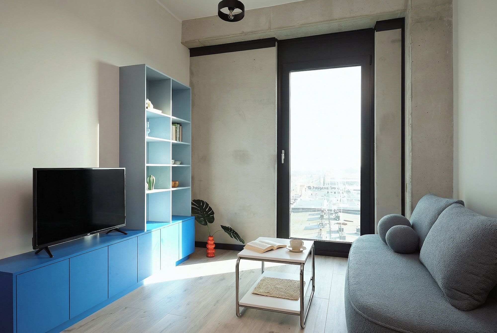 Sala de estar com televisão, estantes azuis, uma mesinha com um livro e uma chávena, e um sofá cinzento.