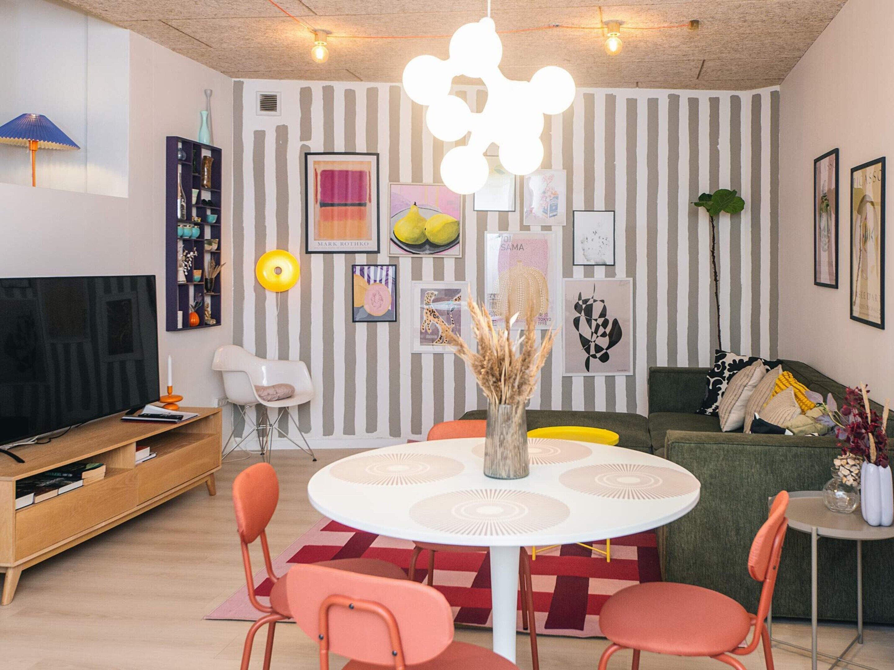 Uma sala de estar moderna com uma mesa de jantar redonda, cadeiras laranja, um sofá verde e uma TV sobre um suporte de madeira.