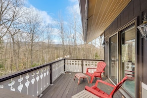 Um deck de madeira com duas cadeiras Adirondack vermelhas e uma pequena mesa, com vista para uma área arborizada.