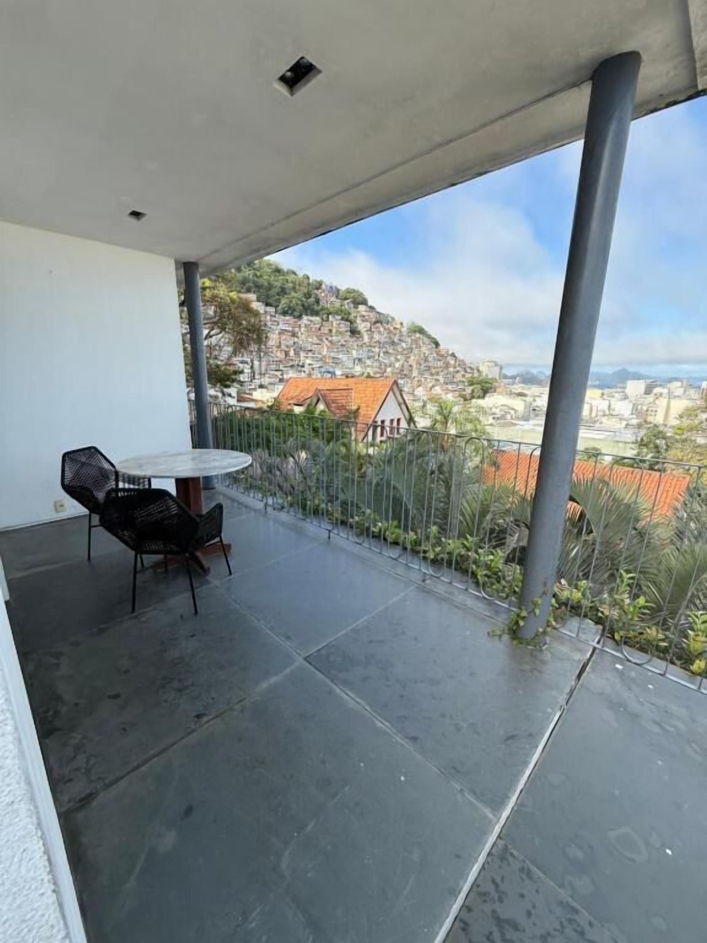 Uma varanda com mesa e cadeiras, vista para uma cidade no alto de uma colina e grade de metal.