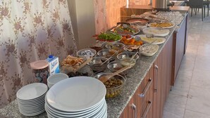 Disposição de buffet com diversos alimentos, um bebedouro e uma pilha de pratos.