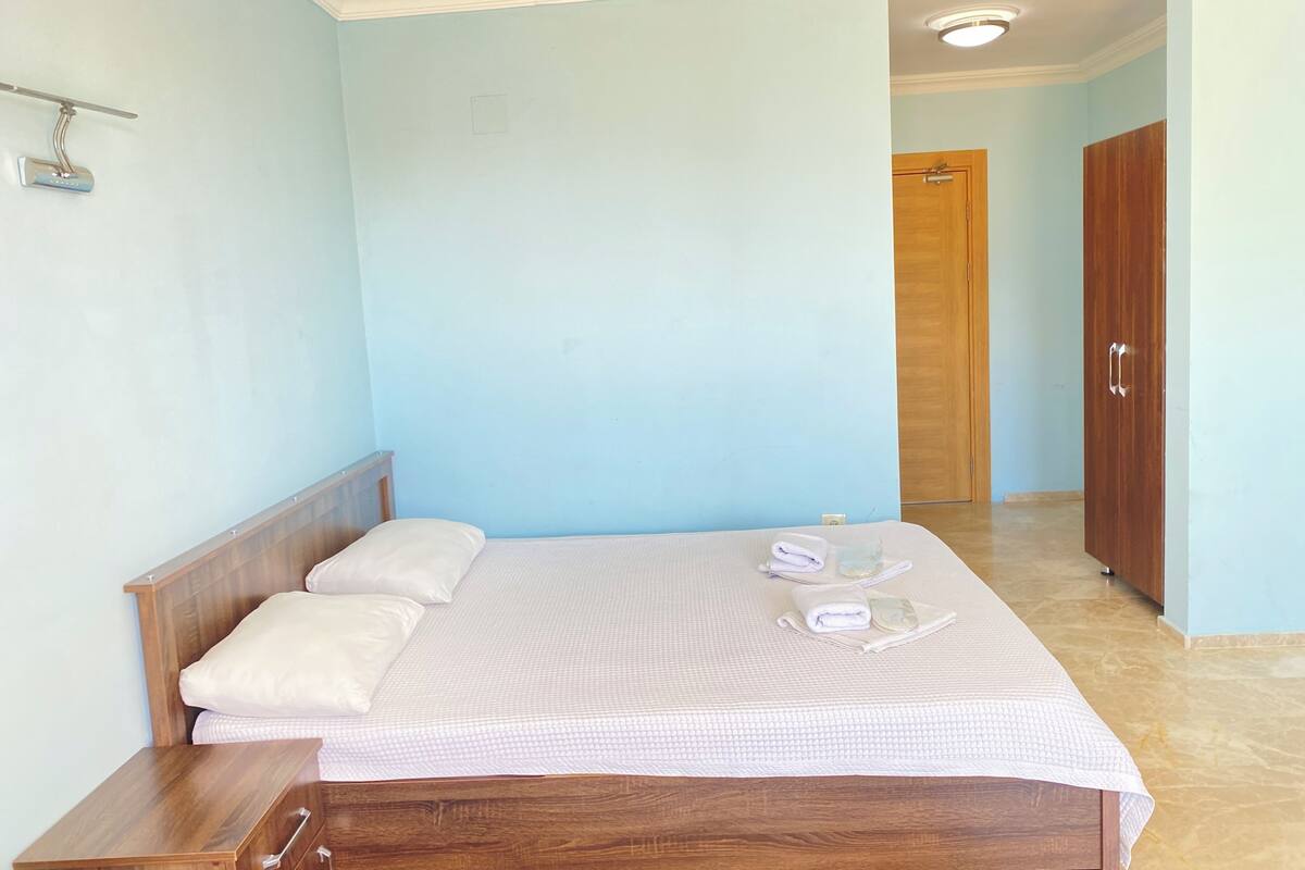 Quarto com cama, cabeceira de madeira, mesa de cabeceira, guarda-roupa e banheiro com chuveiro.