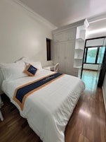 Cama bem arrumada com colcha branca e azul, uma pequena escrivaninha com cadeira e um guarda-roupa com painéis decorativos.