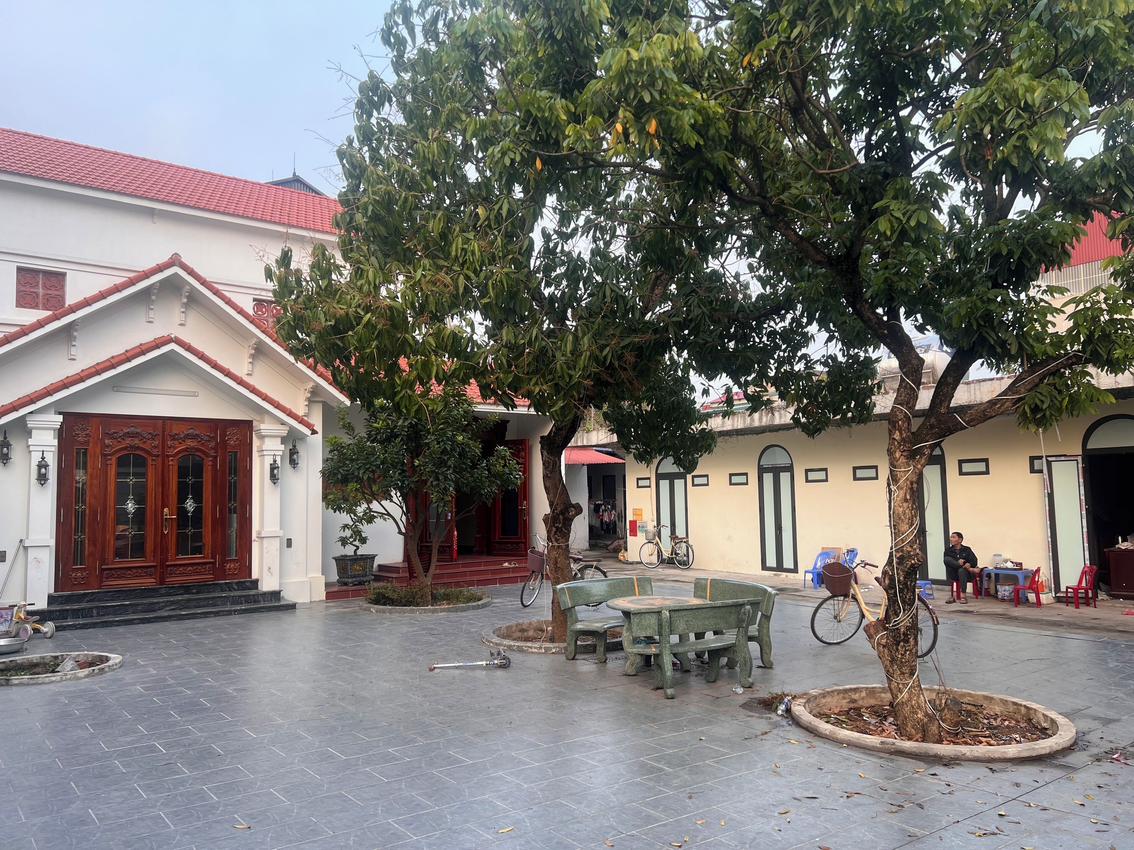 Um pátio com um edifício, apresentando telhado de telhas vermelhas e porta de madeira tradicional. Há áreas de estar ao ar livre com bancos e mesas, cercadas por árvores.