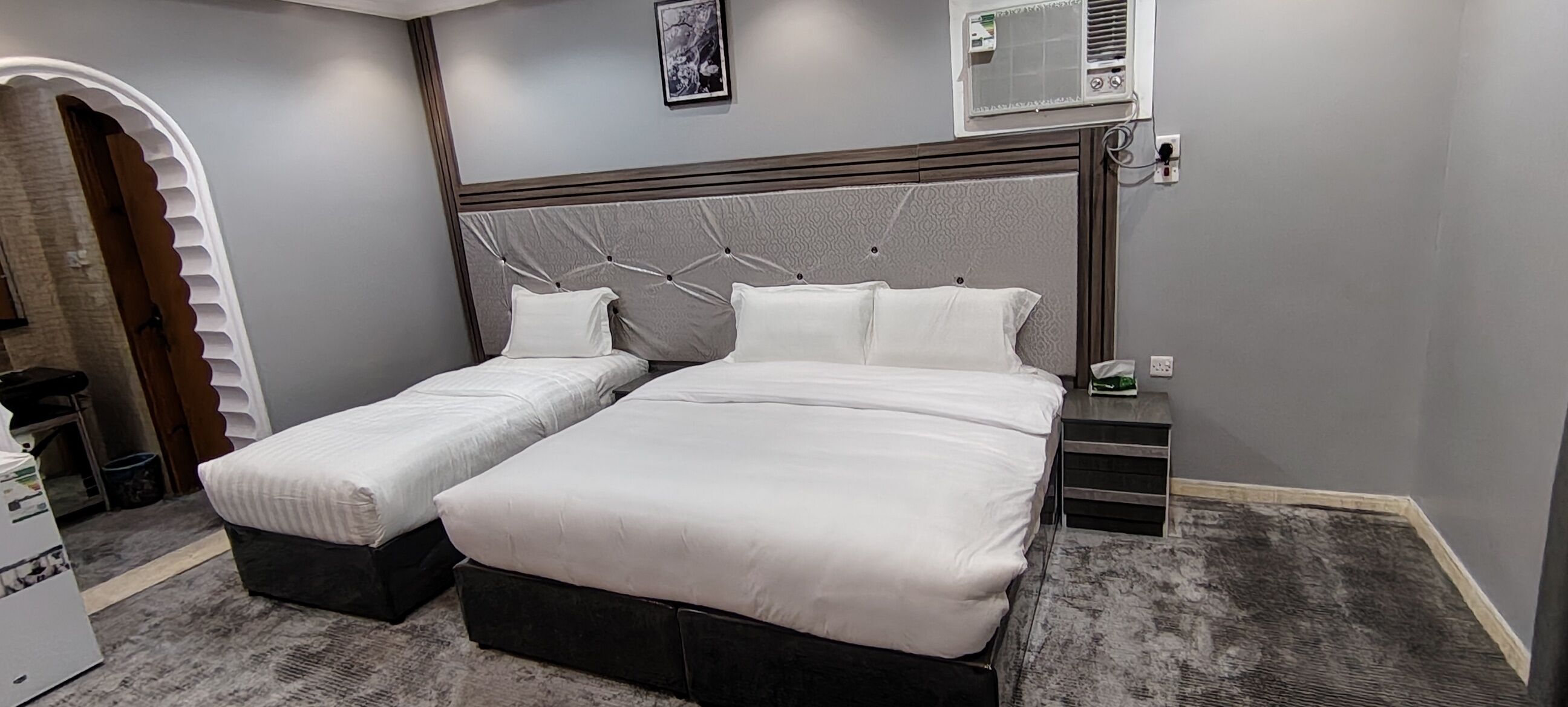 Uma cama de casal com cabeceira, mesa de cabeceira e uma pequena luminária.