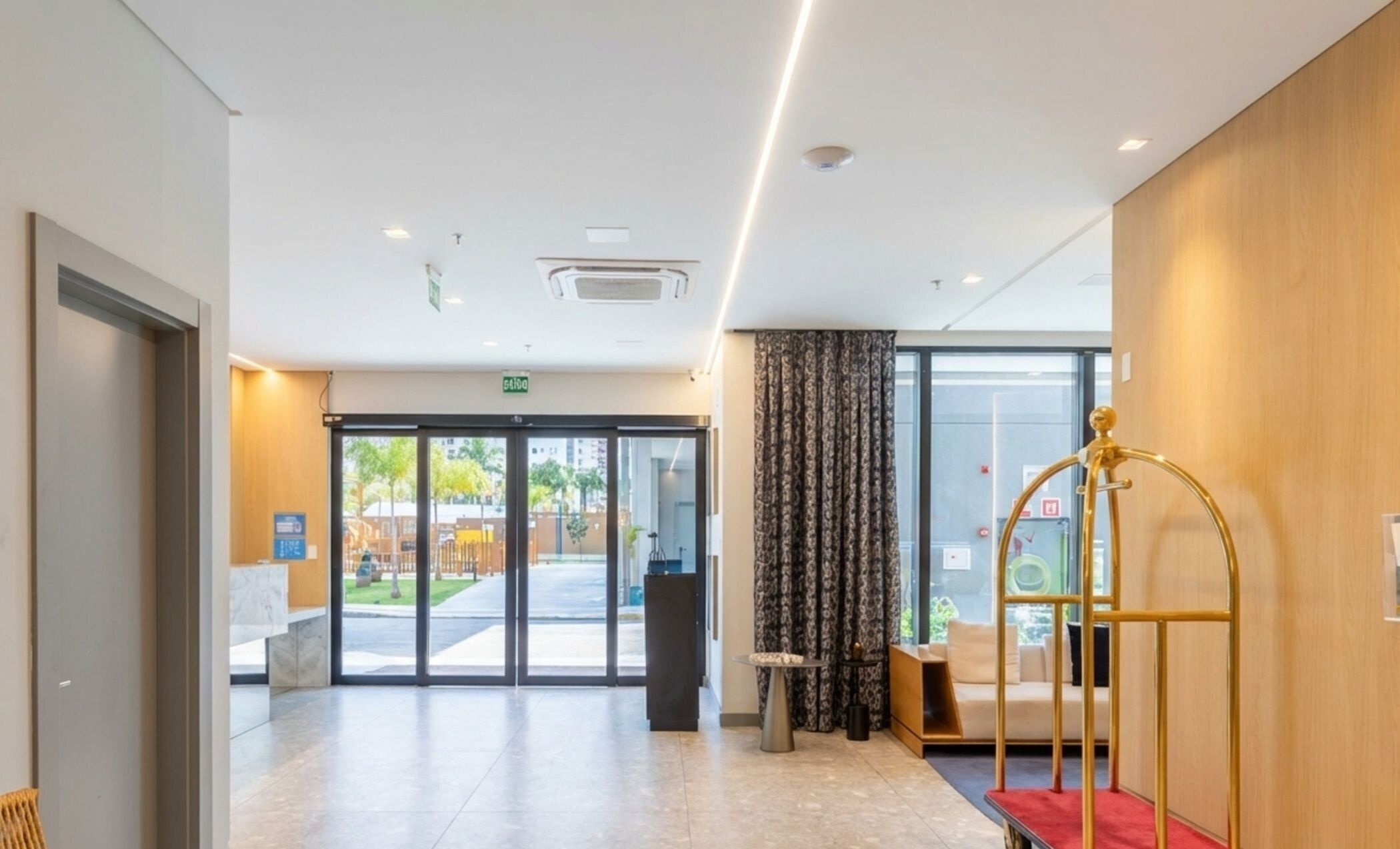 Um saguão de hotel com uma mesa de recepção, área de estar com sofá e poltronas, uma janela ampla com vista para o exterior e um tapete vermelho que leva à entrada.