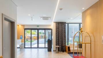 Um saguão de hotel com uma mesa de recepção, área de estar com sofá e poltronas, uma janela ampla com vista para o exterior e um tapete vermelho que leva à entrada.