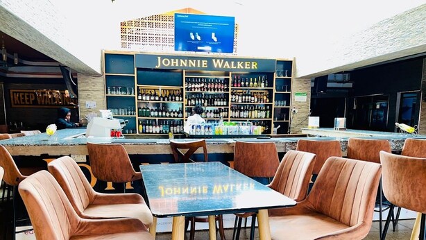 Uma área de bar com um letreiro indicando 'JOHNIE WALKER' e uma variedade de garrafas de bebidas. Há várias cadeiras estofadas e uma mesa em primeiro plano.