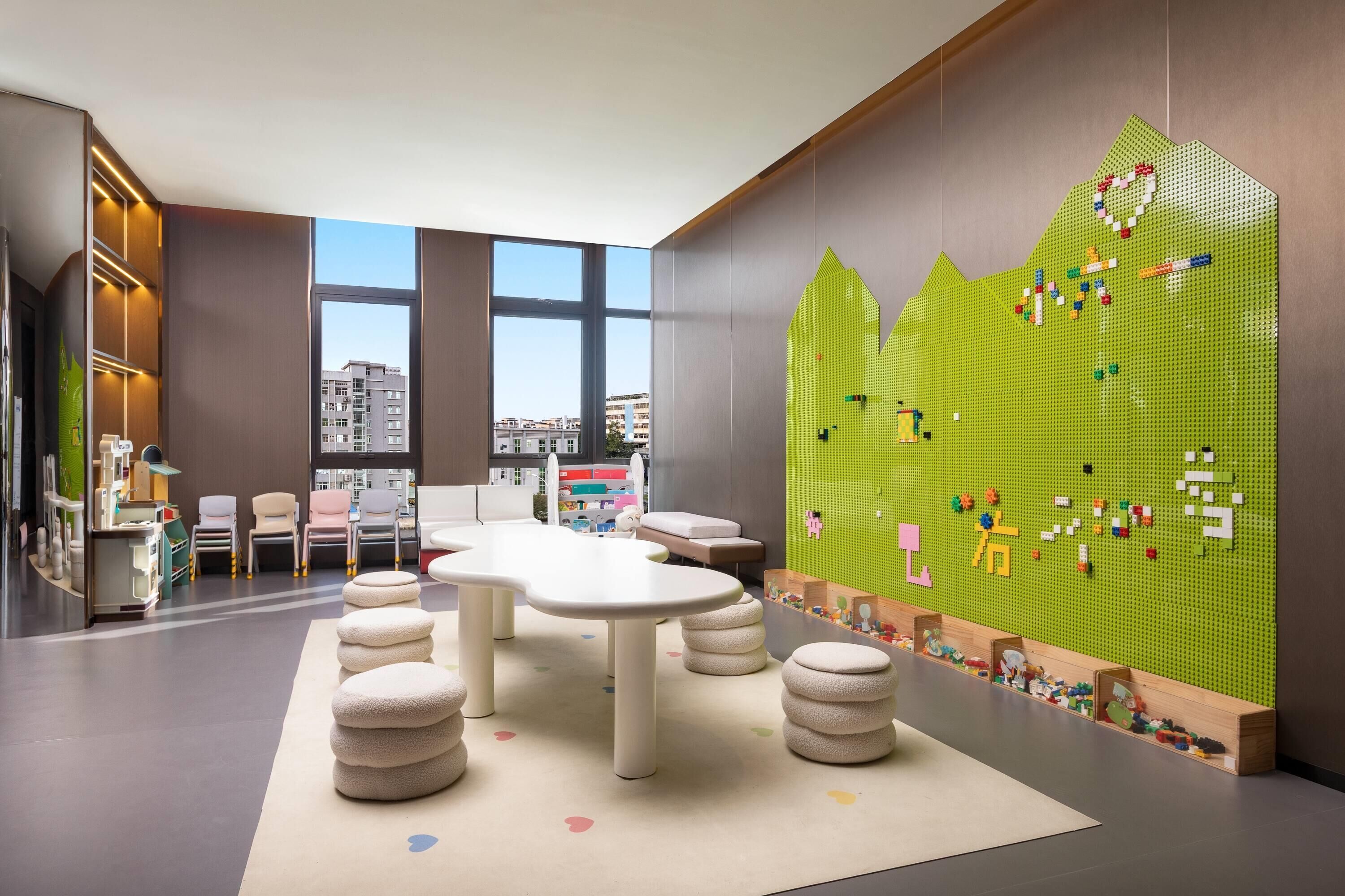 Uma área de recreação infantil moderna, com uma mesa grande e assentos, uma parede de blocos LEGO e vista para edifícios.