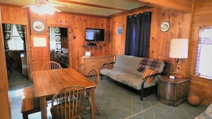 Interior de uma cabana de madeira com mesa de jantar, cadeiras, sofá, televisão e um abajur.