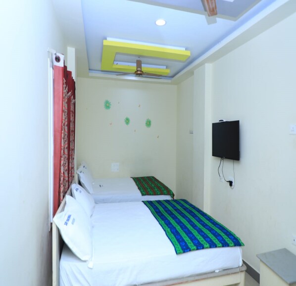 Quarto pequeno com duas camas, um ventilador de teto, uma TV fixada na parede e uma janela com cortinas.