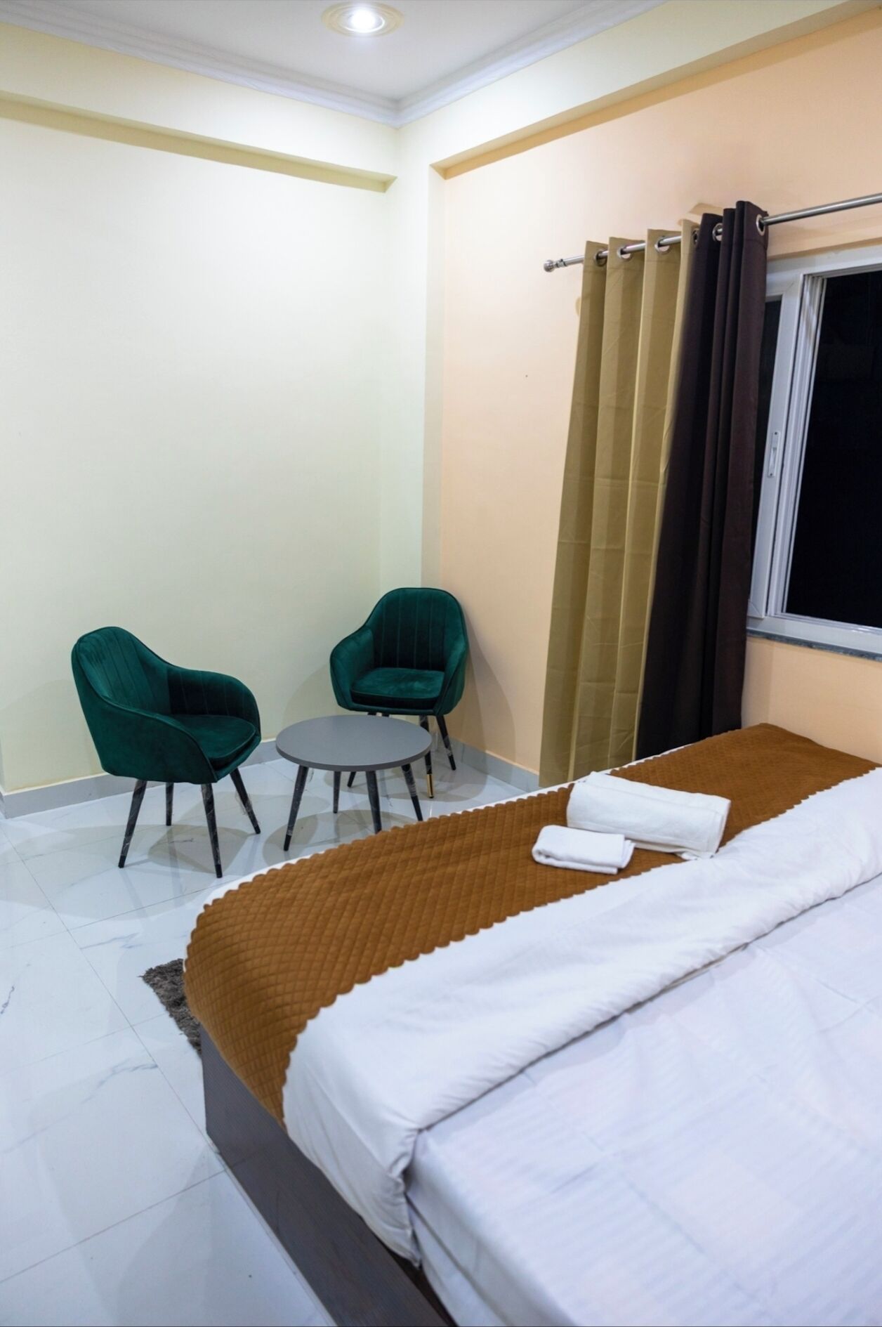Quarto de hotel com uma cama, duas cadeiras verdes, uma mesinha redonda e uma janela com cortinas.