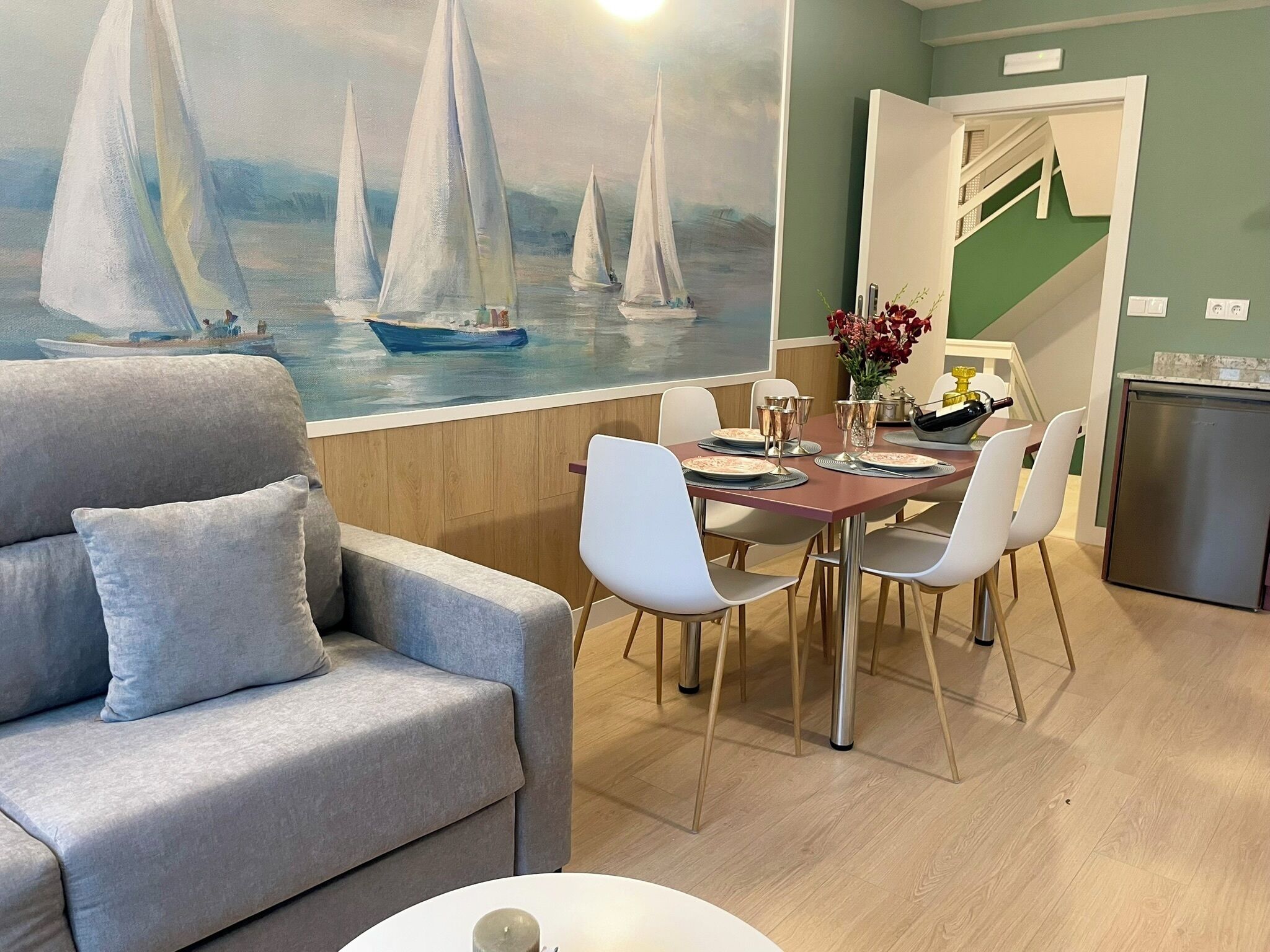 Uma sala de estar moderna com um sofá cinzento, mesa de jantar com cadeiras e um quadro de veleiros na parede.