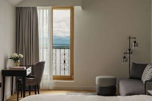 Quarto de hotel com cama, escrivaninha com cadeira, um pequeno banquinho, mesa de cabeceira e vista para montanhas pela janela.
