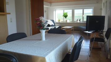 Uma sala de jantar com uma mesa posta para quatro pessoas, um sofá e uma televisão ao fundo.