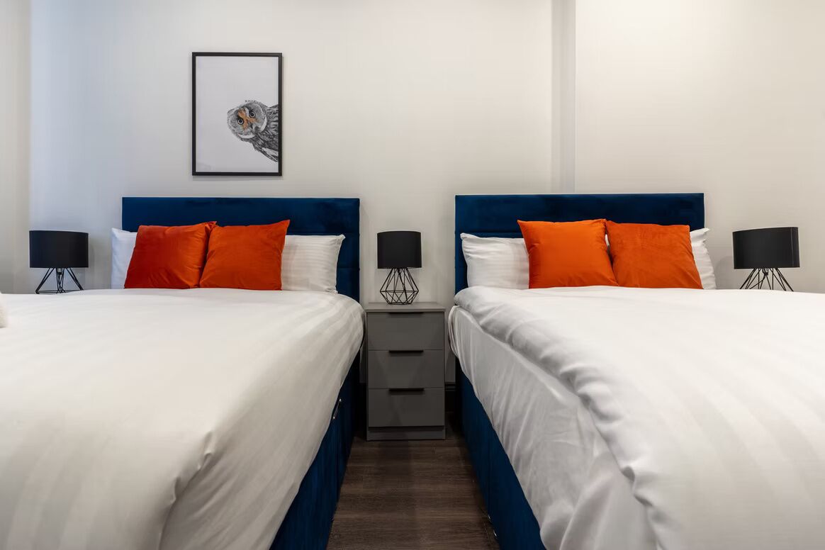 Quarto de hotel com duas camas, roupa de cama branca, almofadas laranja e abajures pretos.