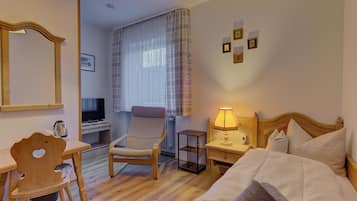 Quarto de hotel com cama, escrivaninha e cadeira, televisão, luminária e janela com cortinas.