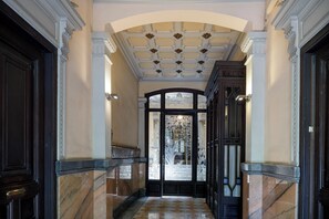 Um amplo salão com piso de mármore, teto ricamente decorado e uma grande porta de vidro que dá para um pátio.