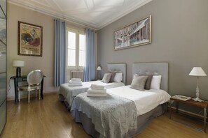 Quarto de hotel com duas camas, uma escrivaninha, uma cadeira, um radiador e uma janela com cortinas.