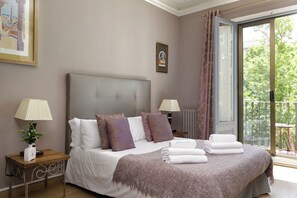 Quarto com cama, duas mesas de cabeceira, janela com cortinas e vista para o verde lá fora.