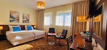 Quarto de hotel com cama, duas cadeiras, uma mesa, televisão e obras de arte nas paredes.