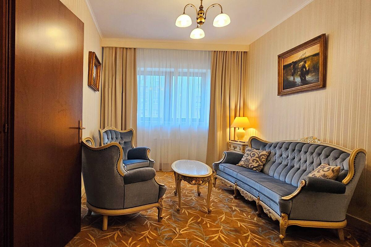 Quarto de hotel com uma cama, duas poltronas, um sofá, uma mesinha e uma janela grande com cortinas.