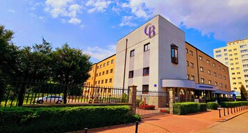 Um edifício moderno com um logotipo 'B' bem visível, entrada com portão e área de estacionamento.