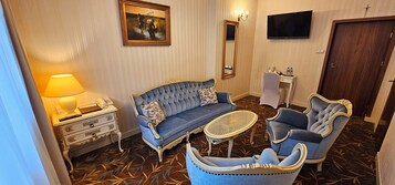 Quarto com sofá azul, poltrona combinando, mesa redonda pequena, mesa de cabeceira com abajur, cômoda, espelho, televisão e porta.