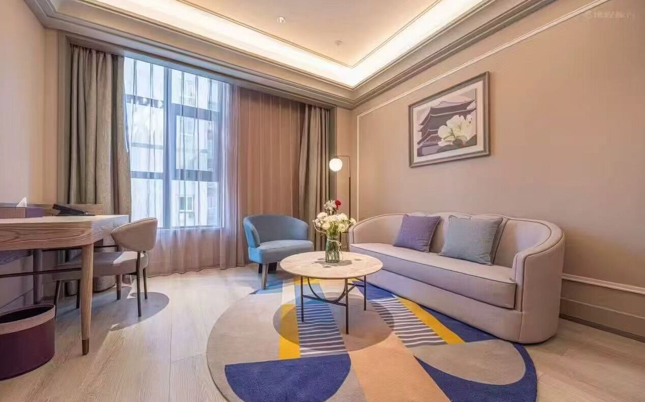 Quarto de hotel moderno com sofá, mesa de jantar e uma cadeira. Há uma janela grande com cortinas, um abajur e um quadro na parede.