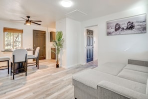 Uma sala de estar moderna com um sofá cinzento, um espaço de jantar com mesa e cadeiras, um ventilador de teto e uma porta que dá para outro quarto.