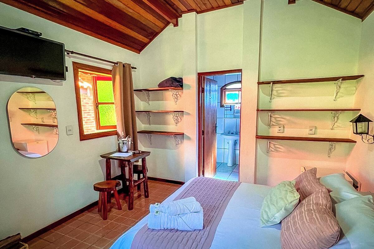 Quarto de hotel com cama, escrivaninha, cadeira, espelho e prateleiras com toalhas.