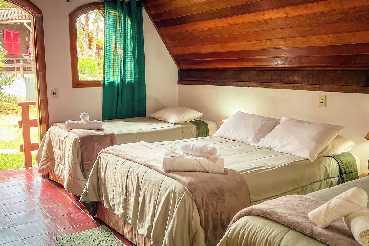 Quarto com duas camas, cada uma com travesseiro e toalha enrolada. Há uma janela com cortinas verdes e teto de madeira.