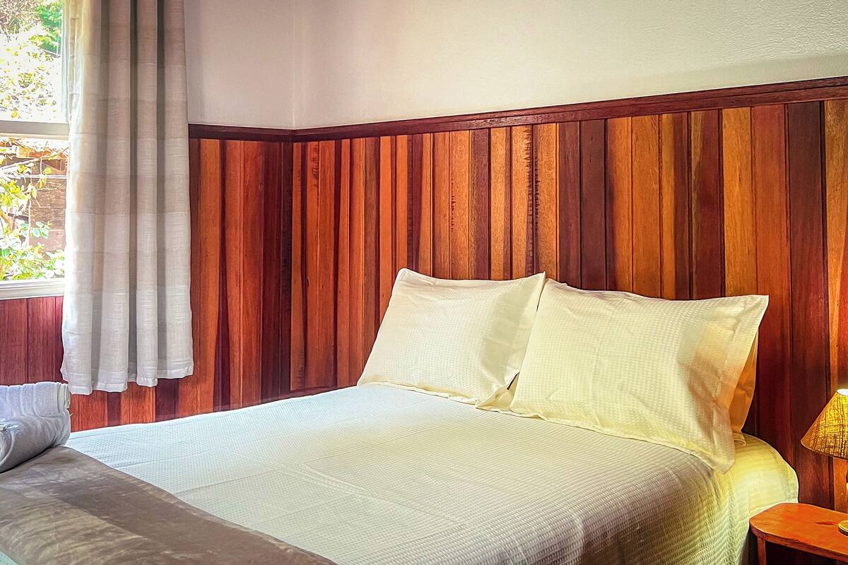 Quarto de hotel com cabeceira em painel de madeira, cama com roupa de cama branca e janela com cortinas.