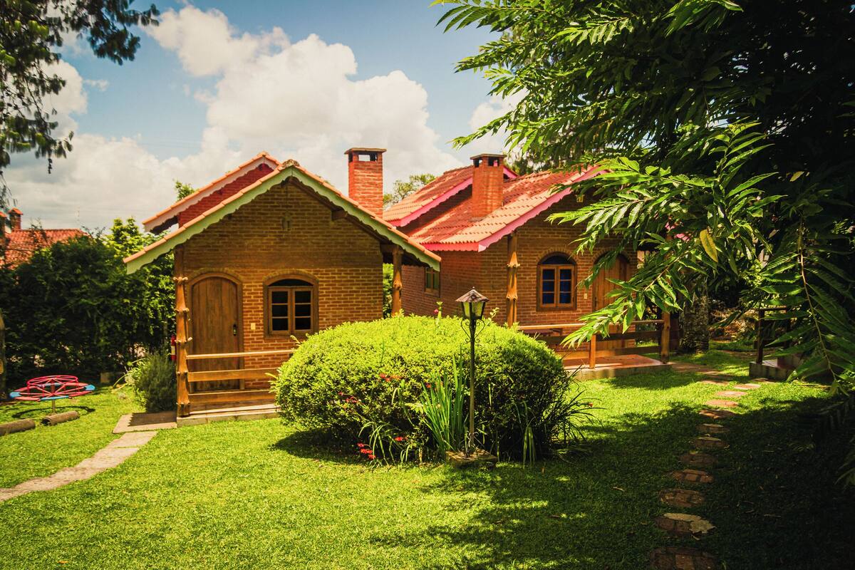 Uma casa de tijolos com telhado vermelho, rodeada por um jardim bem cuidado e um céu limpo.