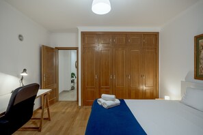 Quarto com cama, escrivaninha, cadeira, guarda-roupa e porta que dá para outro espaço.