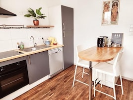 Cozinha compacta com pia, fogão e armários, uma mesa de jantar de madeira com cadeiras e um vaso de planta na bancada.