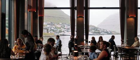 Restaurante com grandes janelas dando para a água e uma cidade ao longe.