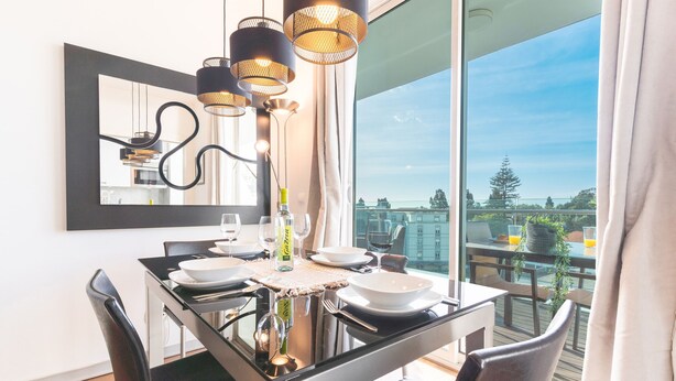 Uma área de refeições moderna com mesa de vidro, cadeiras pretas e luminárias pendentes. Um espelho grande e uma vista da cidade são visíveis.